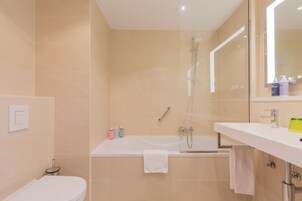 Um banheiro moderno com banheira, chuveiro e vaso sanitário. Há produtos de higiene no lavatório e uma toalha pendurada na parede.