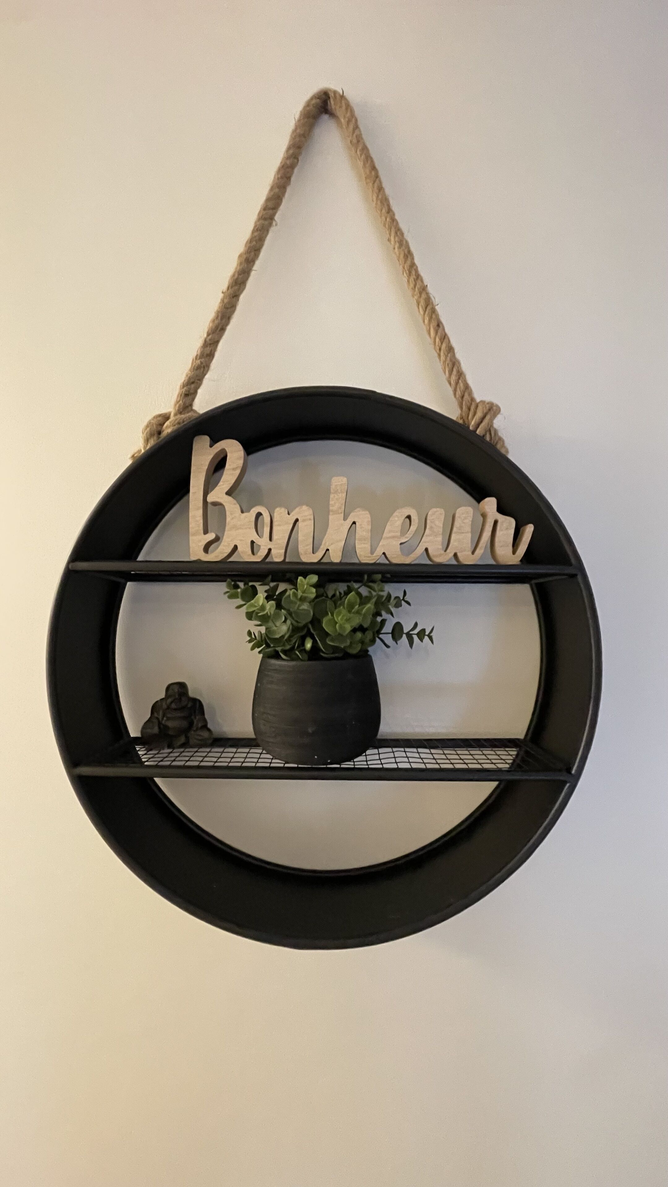 Estante circular presa à parede com a palavra "Bonjour" em fonte decorativa, uma pequena planta em vaso e uma figurina.