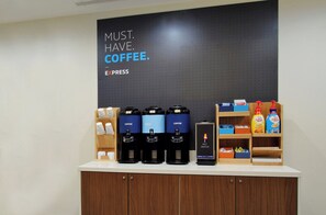 Estação de café com diversos distribuidores de café, um dispensador de água quente e uma placa com a inscrição 'COFÉ ESSENCIAL'.