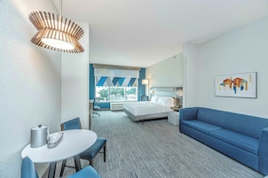 Quarto de hotel com cama, sofá azul, mesa de jantar com cadeiras, abajur e pintura na parede.