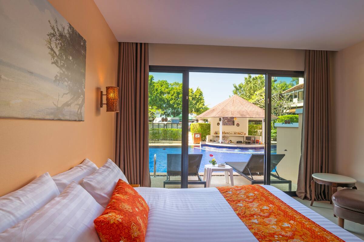 Quarto de hotel com cama, vista para uma piscina e varanda com mesa e cadeiras.