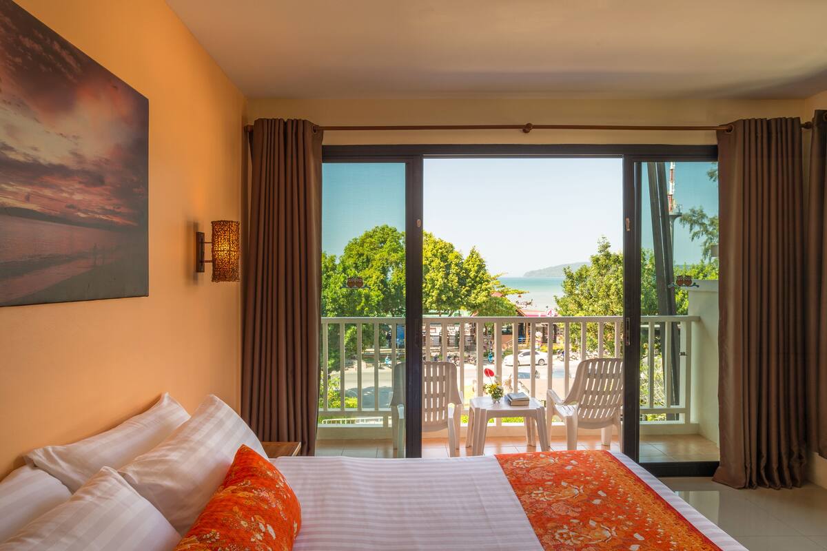 Quarto de hotel com uma cama, um quadro na parede, uma varanda com vista para o mar e uma mesa com cadeiras.
