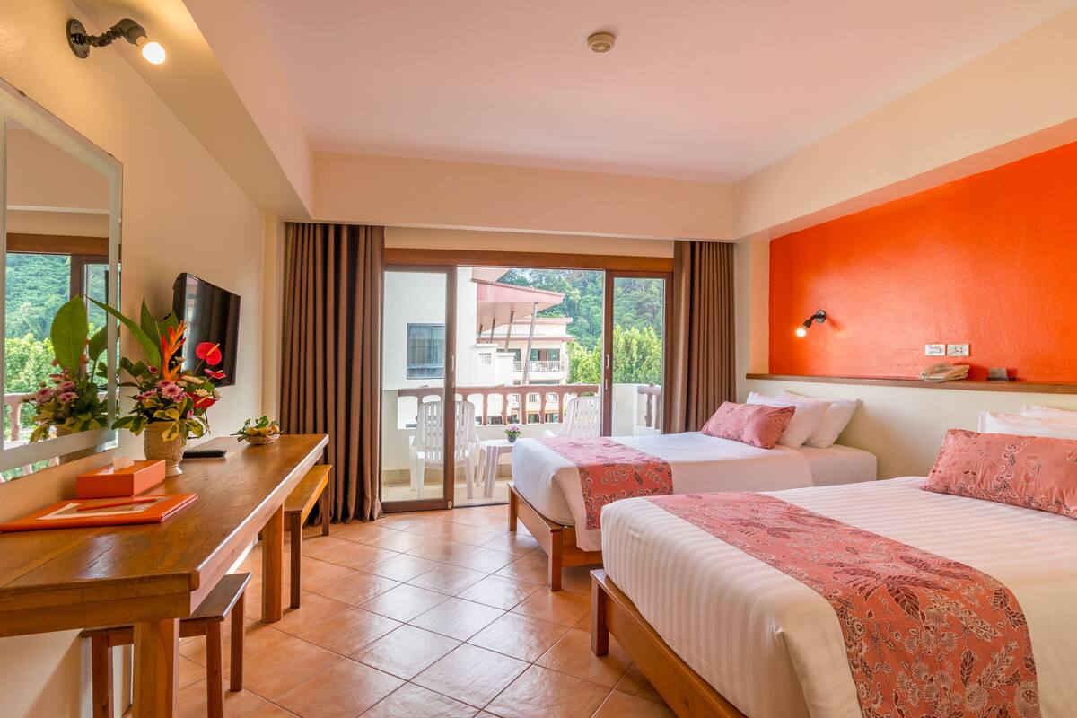 Quarto de hotel com duas camas, uma escrivaninha de madeira, televisão e varanda com vista para o verde.