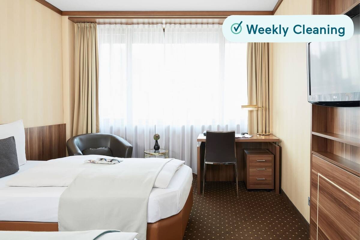 Quarto de hotel com cama, escrivaninha, cadeira, televisão e janela com cortinas.