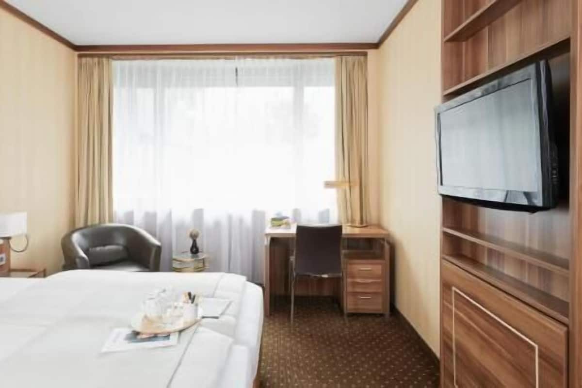 Quarto de hotel com cama, escrivaninha, cadeira, televisão e janela com cortinas.