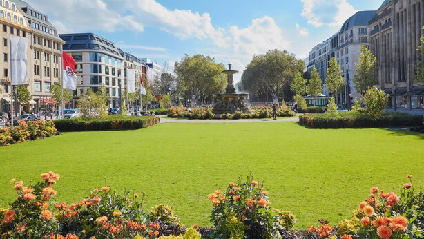 Uma praça pública bem cuidada, com uma fonte central, rodeada por relvado aparado e canteiros de flores.