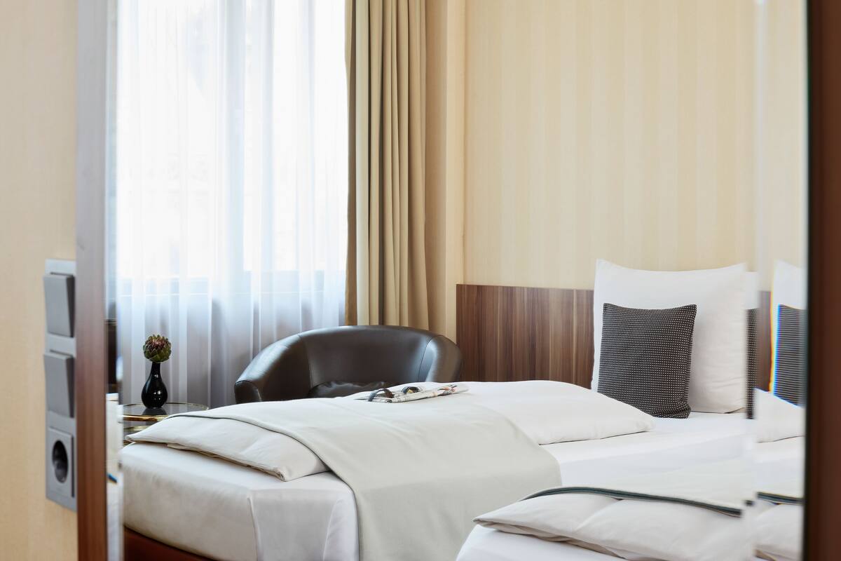 Quarto de hotel com cama, uma cadeira, mesa de cabeceira e janela com cortinas.