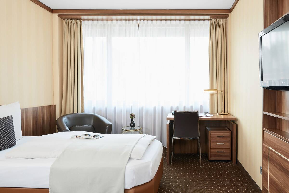 Quarto de hotel com cama, escrivaninha, cadeira, televisão e janela com cortinas.