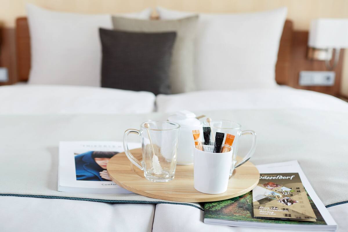 Cama bem arrumada com travesseiro, caneca de vidro com sachê de chá e um livreto sobre um tabuleiro de madeira.