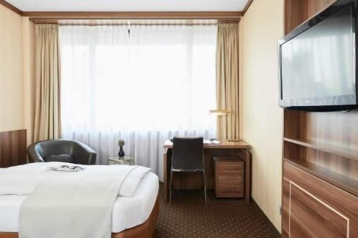 Quarto de hotel com cama, escrivaninha, cadeira, televisão e janela com cortinas.