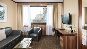 Quarto de hotel com sofá de couro, mesa de centro de vidro com um livro e copos, escrivaninha de madeira com televisão e vista para um edifício histórico através de grandes janelas.