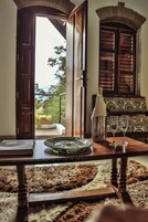 Uma mesa de madeira com um prato decorativo, uma garrafa e dois copos. O quarto possui um sofá estampado e vista para o verde lá fora.