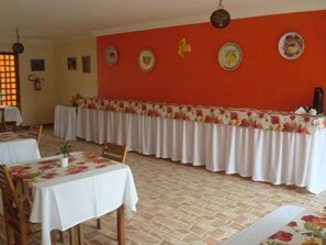 Uma sala de jantar com mesas cobertas por toalhas brancas, piso de tijolos e parede vermelha decorada com pratos.