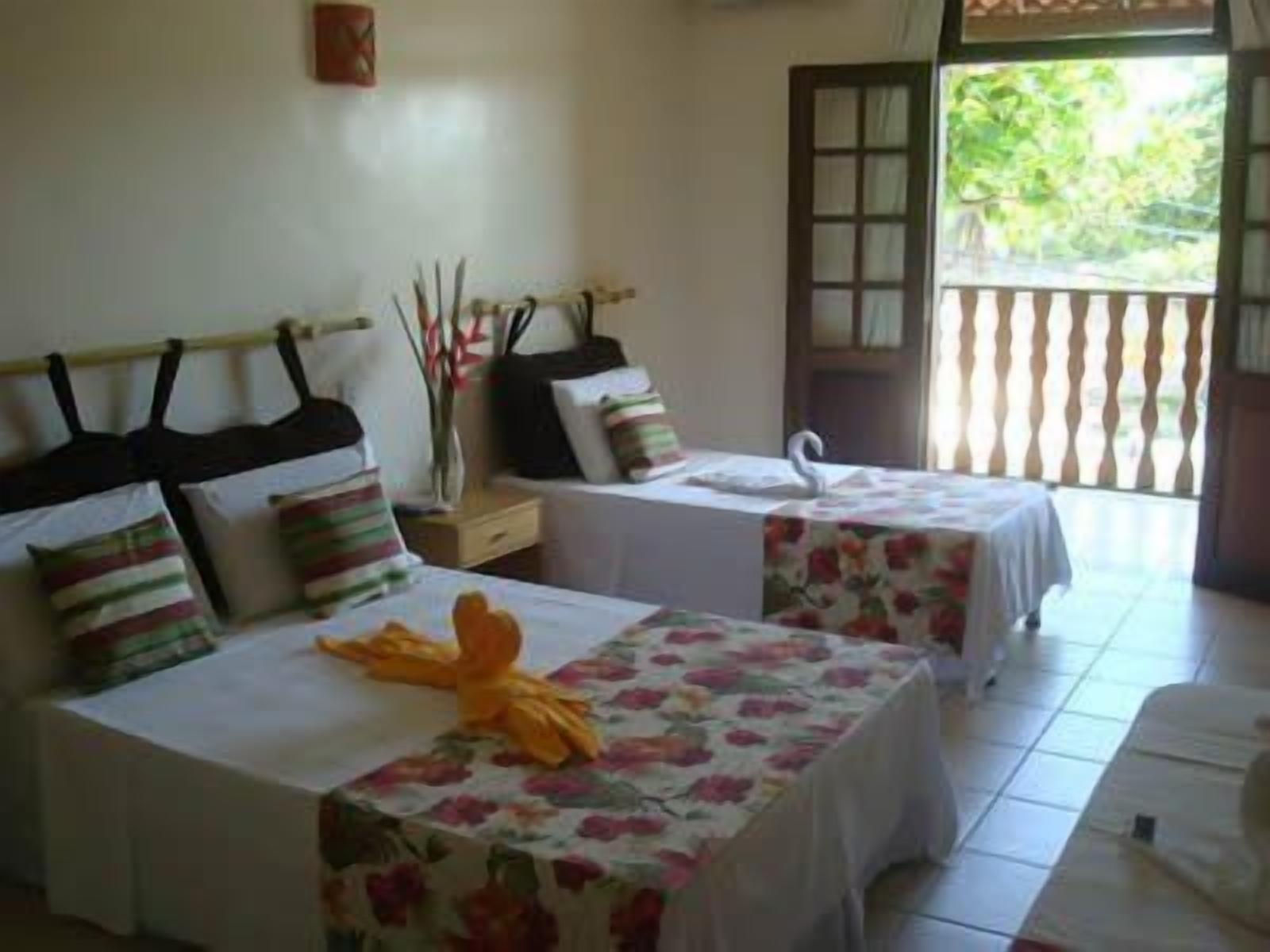 Quarto com duas camas, cada uma com colcha florida e almofadas listradas. Há uma pequena mesa com um vaso de flores e uma decoração de cisne. O quarto possui uma varanda com corrimão de madeira e vista para o verde lá fora.