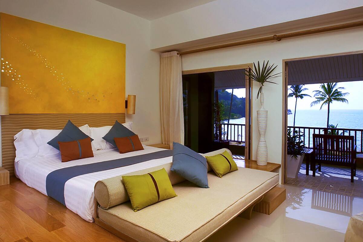 Quarto de hotel com cama, varanda e vista para o oceano, e uma parede amarela grande.