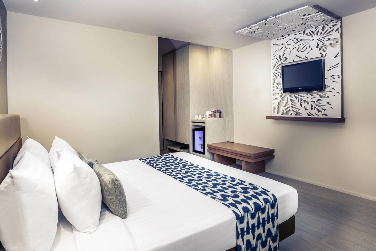 Quarto de hotel com cama, televisão na parede, mesa pequena e painel decorativo.