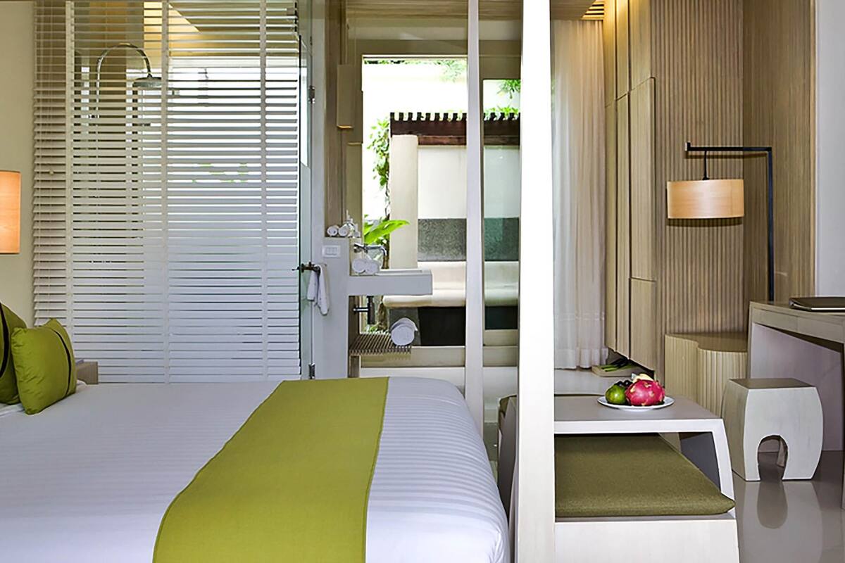 Quarto de hotel moderno com cama, escrivaninha com abajur, espelho e vista para o exterior.