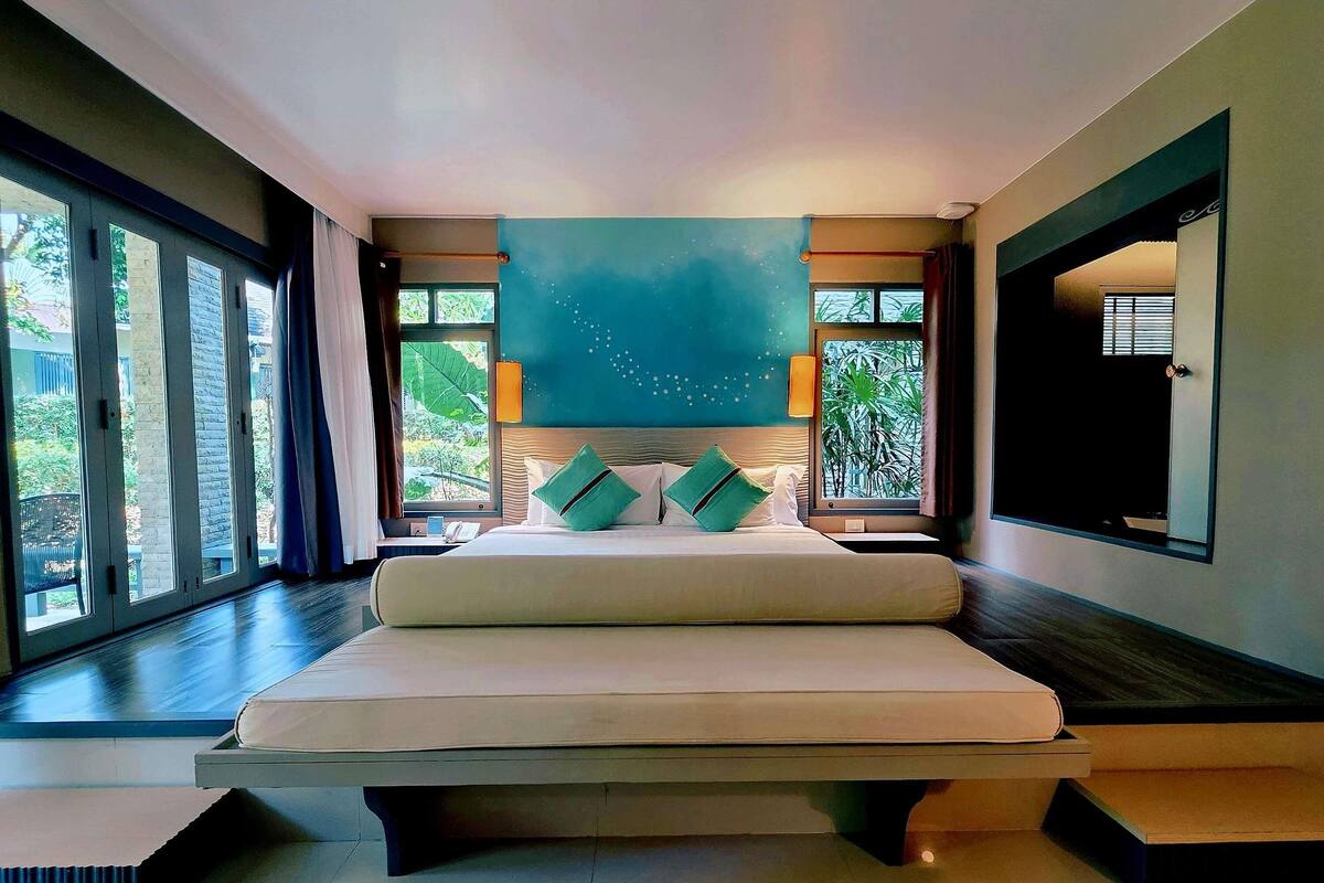 Quarto de hotel moderno com cama grande, duas janelas e vista para área verde.