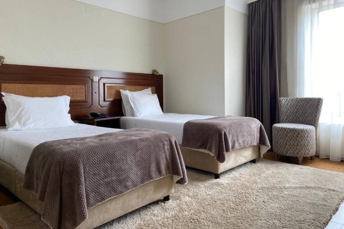 Quarto de hotel com duas camas, cabeceira de madeira, uma poltrona pequena e uma janela grande com cortinas.