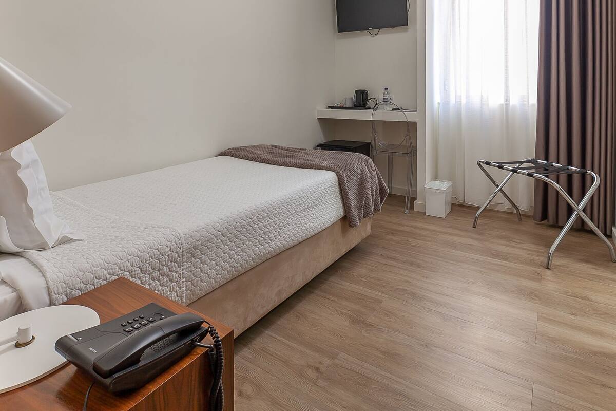 Quarto de hotel com cama, escrivaninha com computador, televisão, abajur e telefone.
