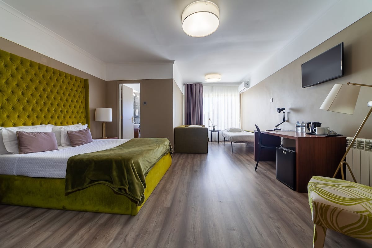 Quarto de hotel com cama grande, escrivaninha, cadeira, televisão e janela com cortinas.