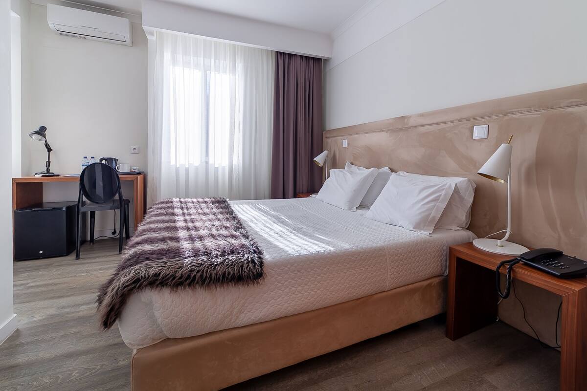 Quarto de hotel com cama, escrivaninha, cadeira, luminária e ar condicionado.