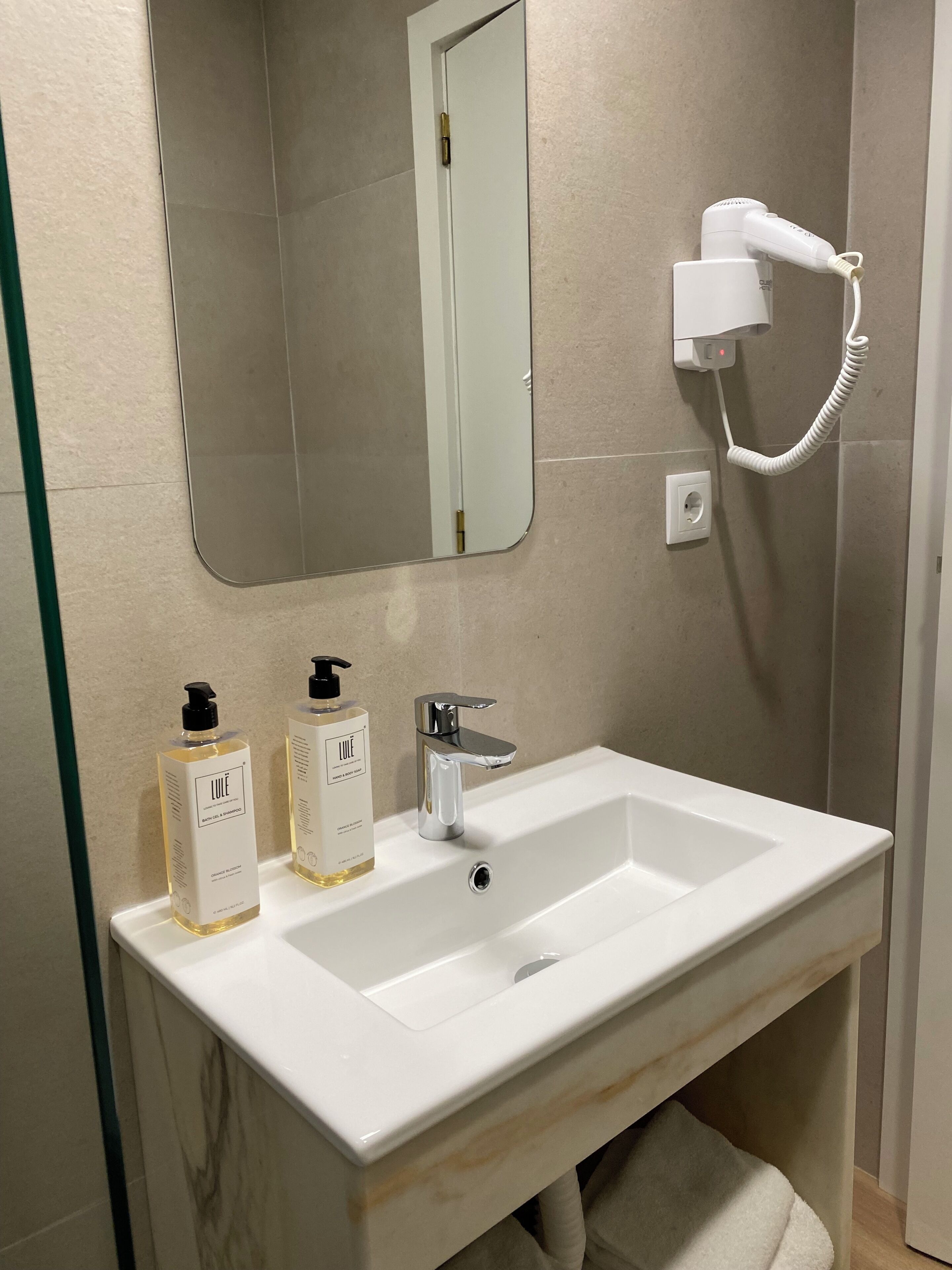 Um banheiro com pia branca, duas garrafas de shampoo LULE, um secador de cabelo e um espelho.
