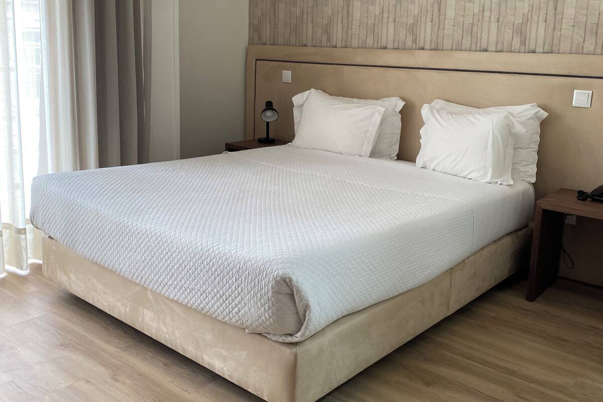 Uma cama grande com cobertura branca acolchoada, cabeceira de madeira e mesa de cabeceira com abajur.