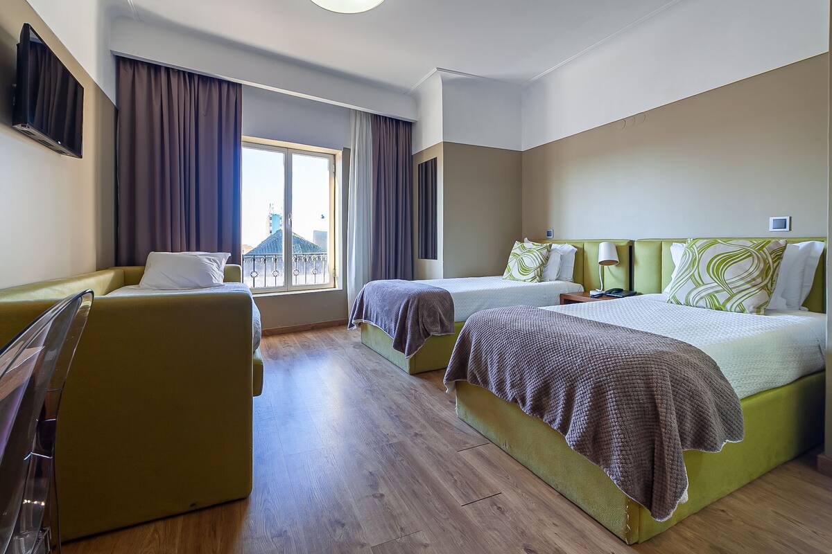 Quarto de hotel com duas camas, um sofá, uma televisão e uma janela com cortinas.