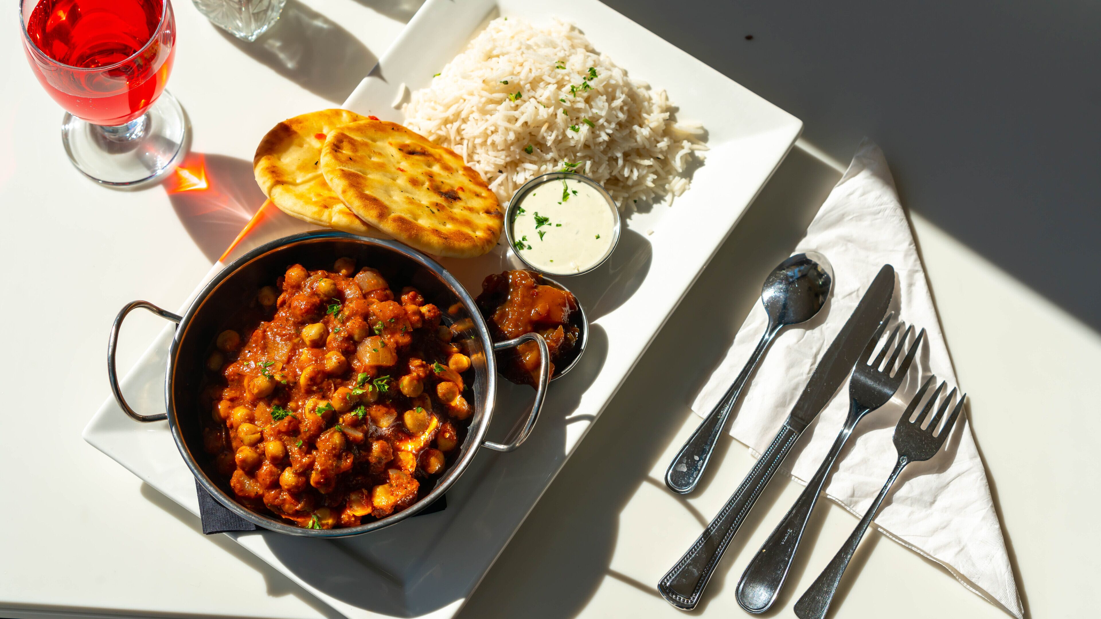 Refeição com arroz, pão naan e ensopado de grão-de-bico, servida num prato branco com talheres e um copo de bebida vermelha.