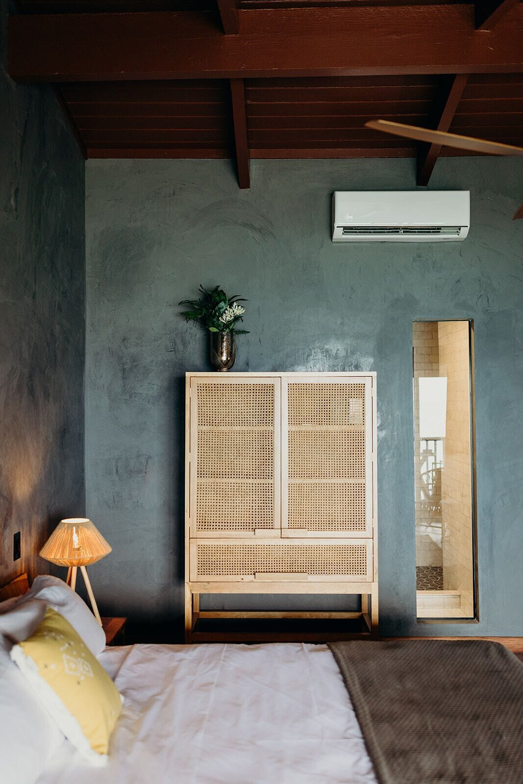 Quarto com cama, luminária de cabeceira, guarda-roupa de rattan, planta em vaso e ar condicionado.