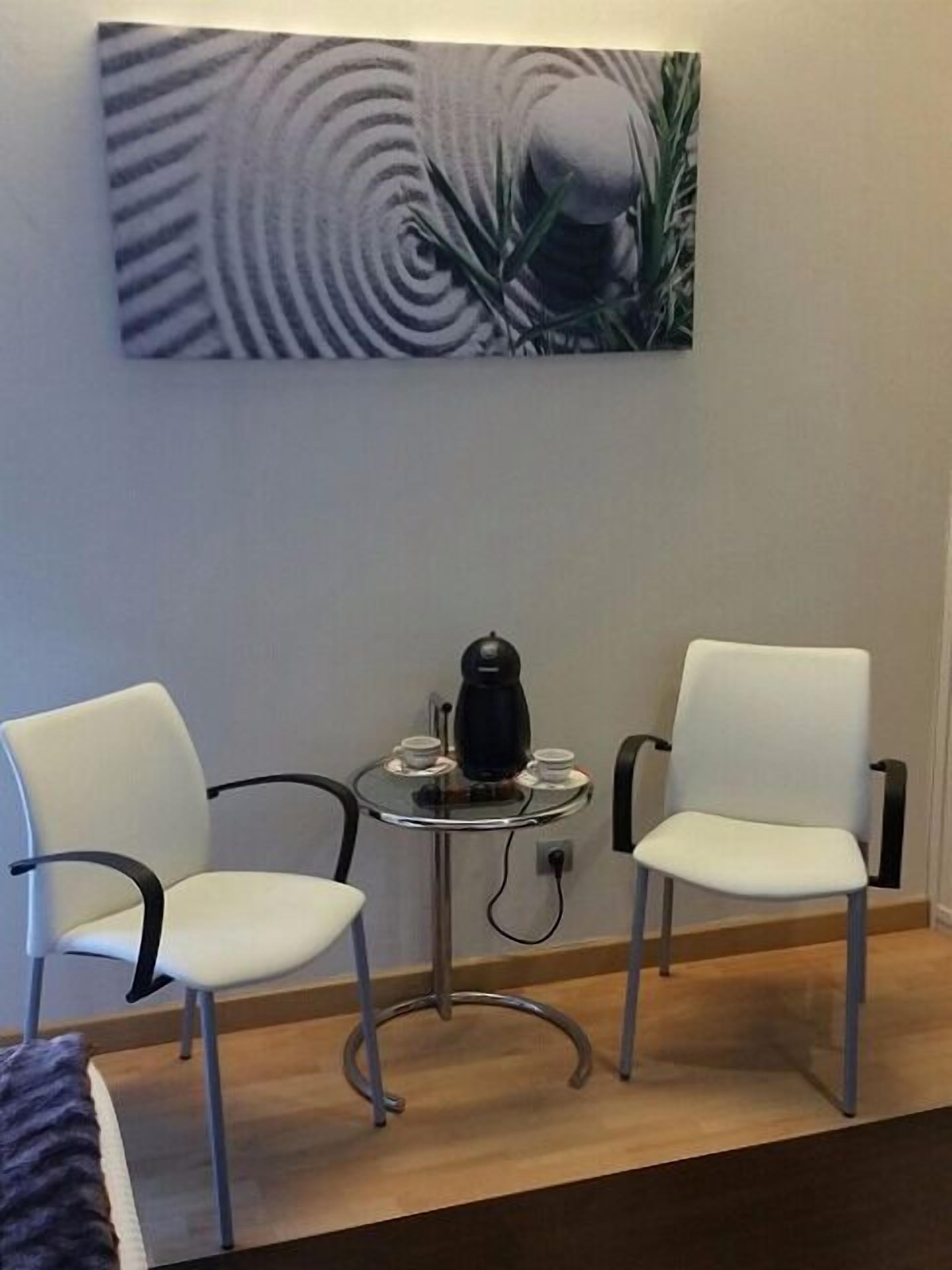 Quarto com duas cadeiras brancas, uma mesa redonda pequena com uma cafeteira preta e uma obra de arte na parede.
