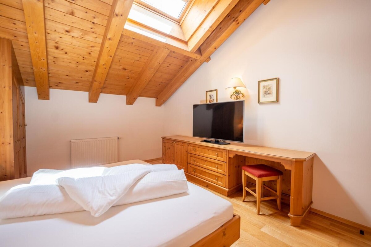 Quarto com teto de madeira, cama, escrivaninha com cadeira, televisão e radiador.