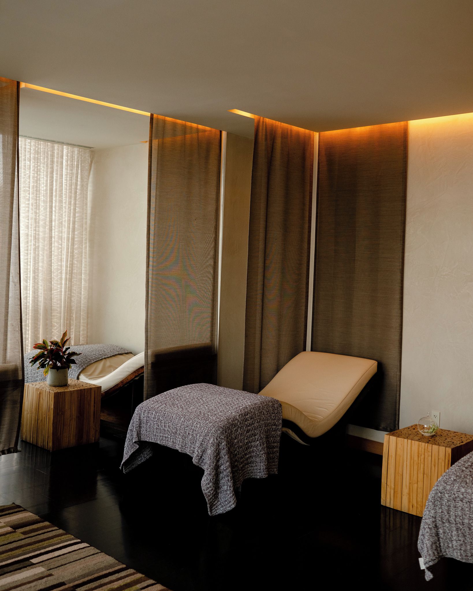 Um quarto de spa com mesa de massagem, uma pequena mesa lateral de madeira com uma planta e um espelho.