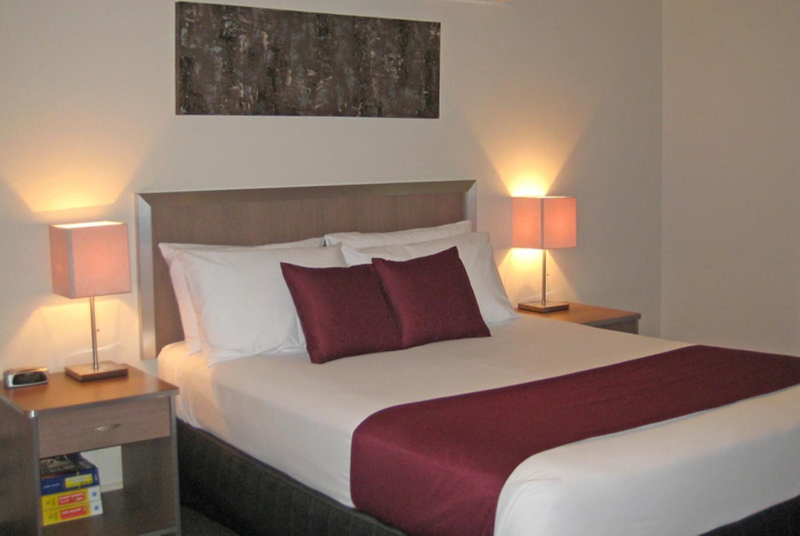 Cama bem arrumada com roupa de cama branca e bordô, duas luminárias de cabeceira e mesa de cabeceira com controle remoto.