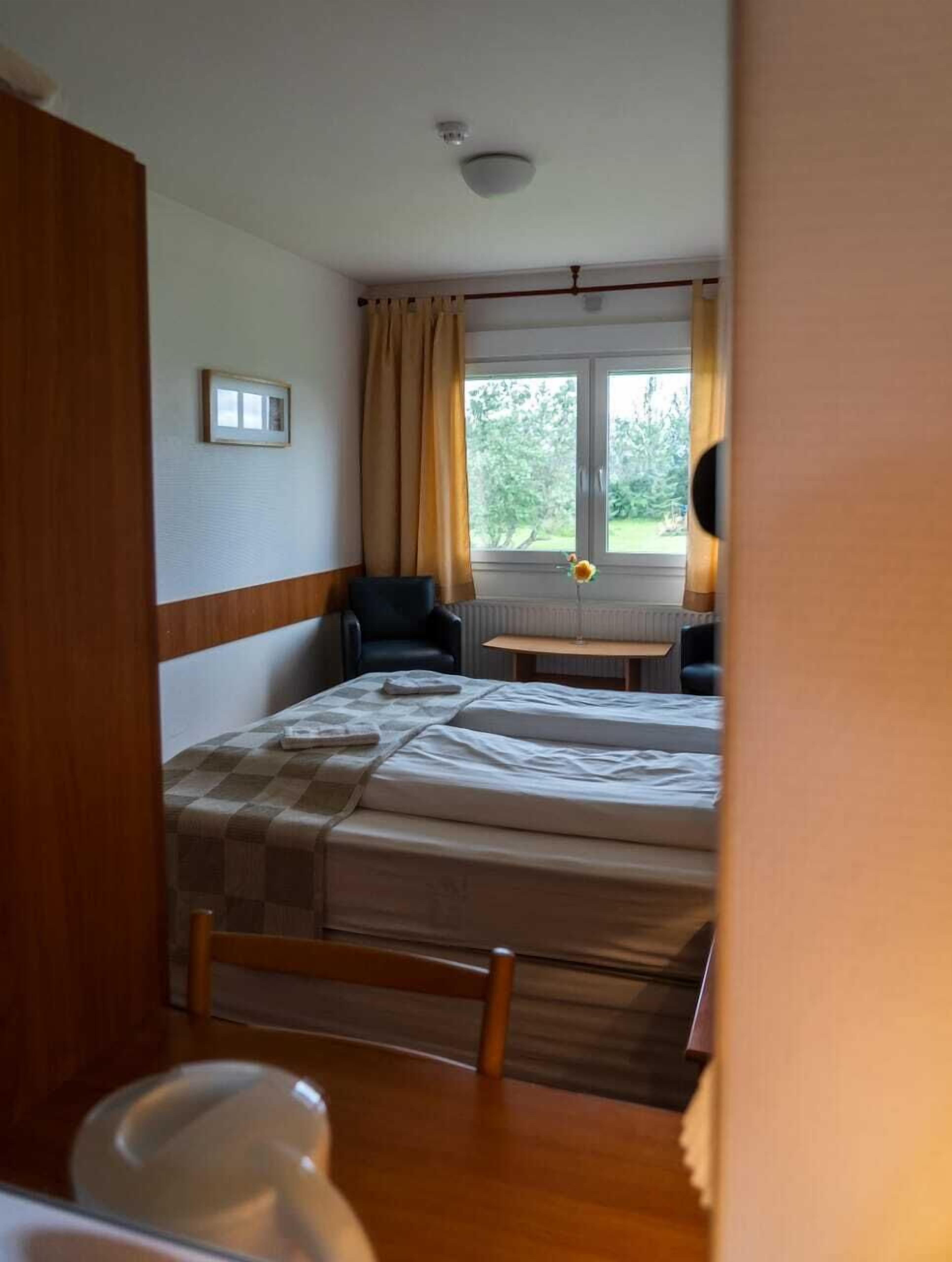 Quarto com cama, uma cadeira, uma mesinha e janela com cortinas.