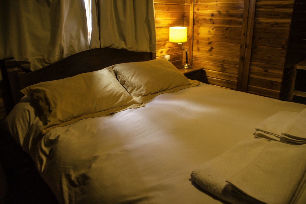 Cama bem arrumada com travesseiros e toalha dobrada, abajur de cabeceira e painéis de madeira nas paredes.