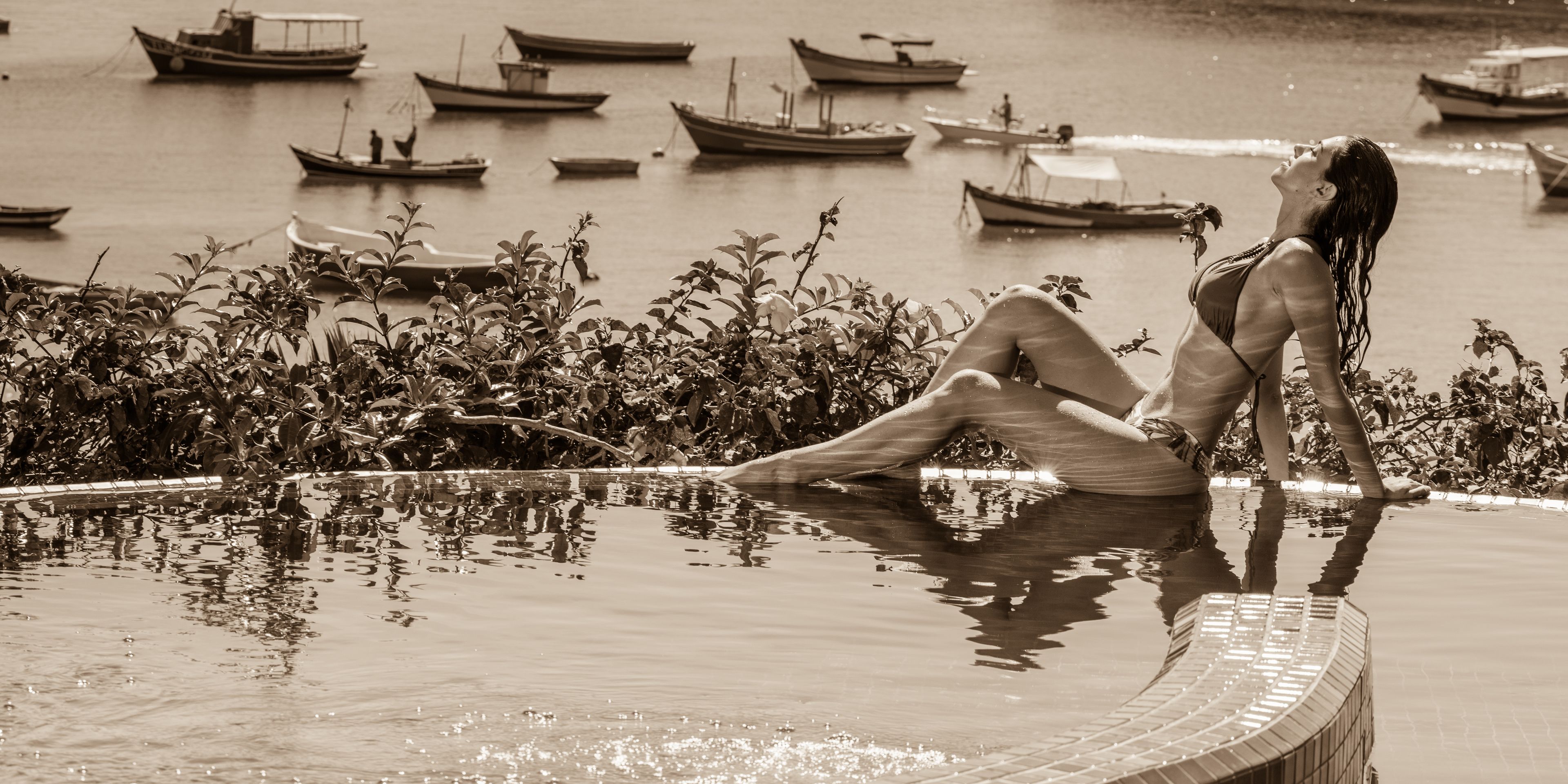 Uma mulher relaxando à beira da piscina, com barcos ao fundo.