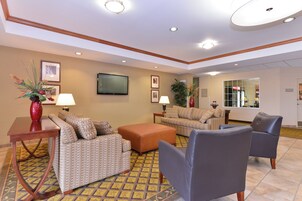 Um saguão de hotel com piso xadrez, sofás, poltronas, uma TV e um balcão de recepção.