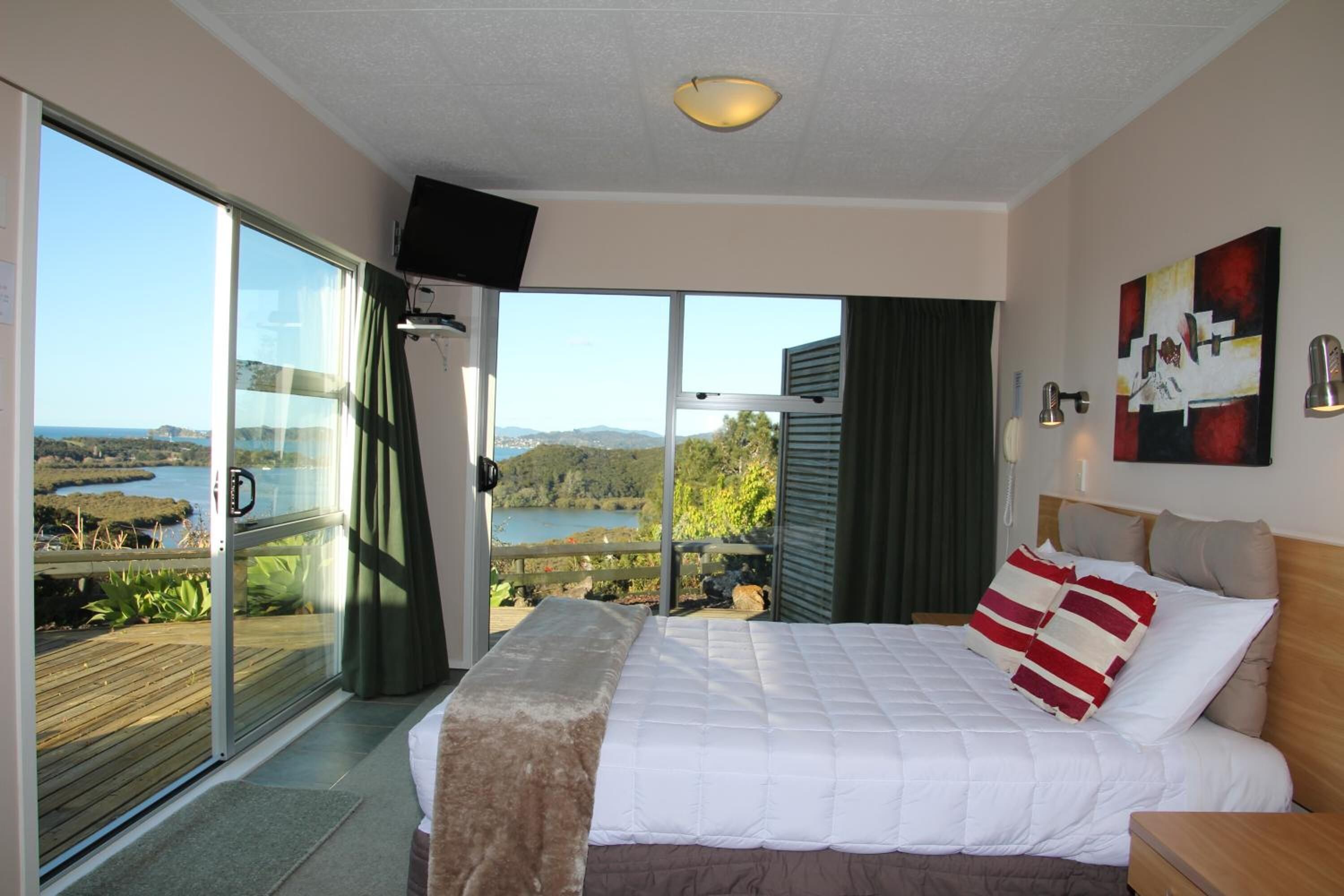 Quarto com uma cama grande, televisão fixada na parede e vista para um lago e colinas.