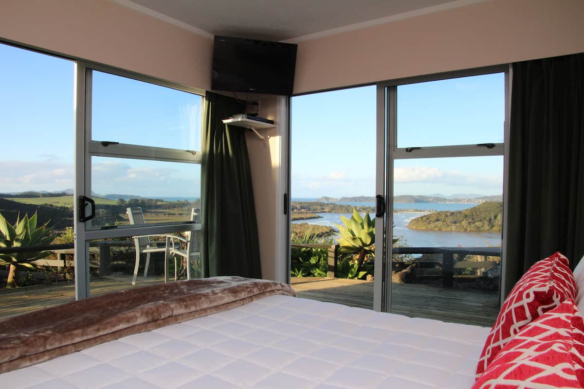 Quarto com uma cama grande, televisão e varanda com vista para um lago e colinas.