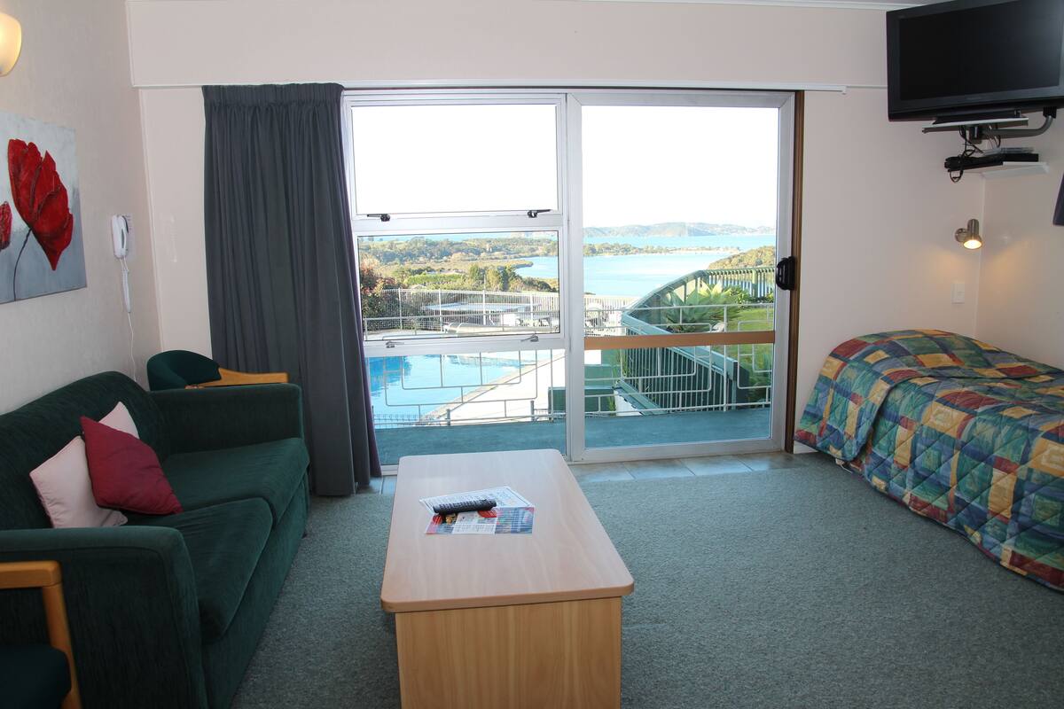 Quarto com sofá verde, mesa de madeira, cama colorida e porta de vidro deslizante que dá para uma varanda com vista para um corpo d'água e uma ponte.
