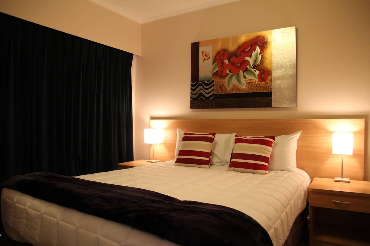 Quarto de hotel com uma cama grande, duas luminárias de cabeceira, um quadro acima da cama e uma cortina escura na janela.