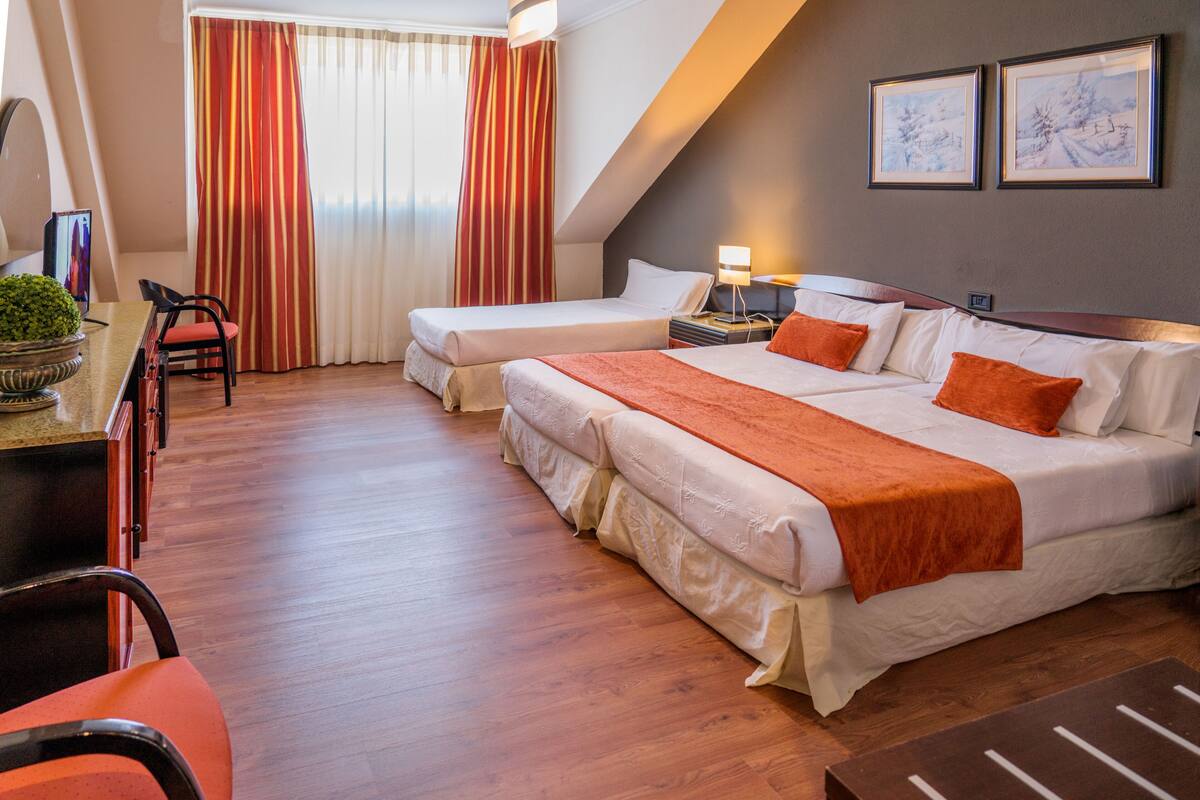 Quarto de hotel com duas camas, uma escrivaninha, uma cadeira e uma janela grande com cortinas.