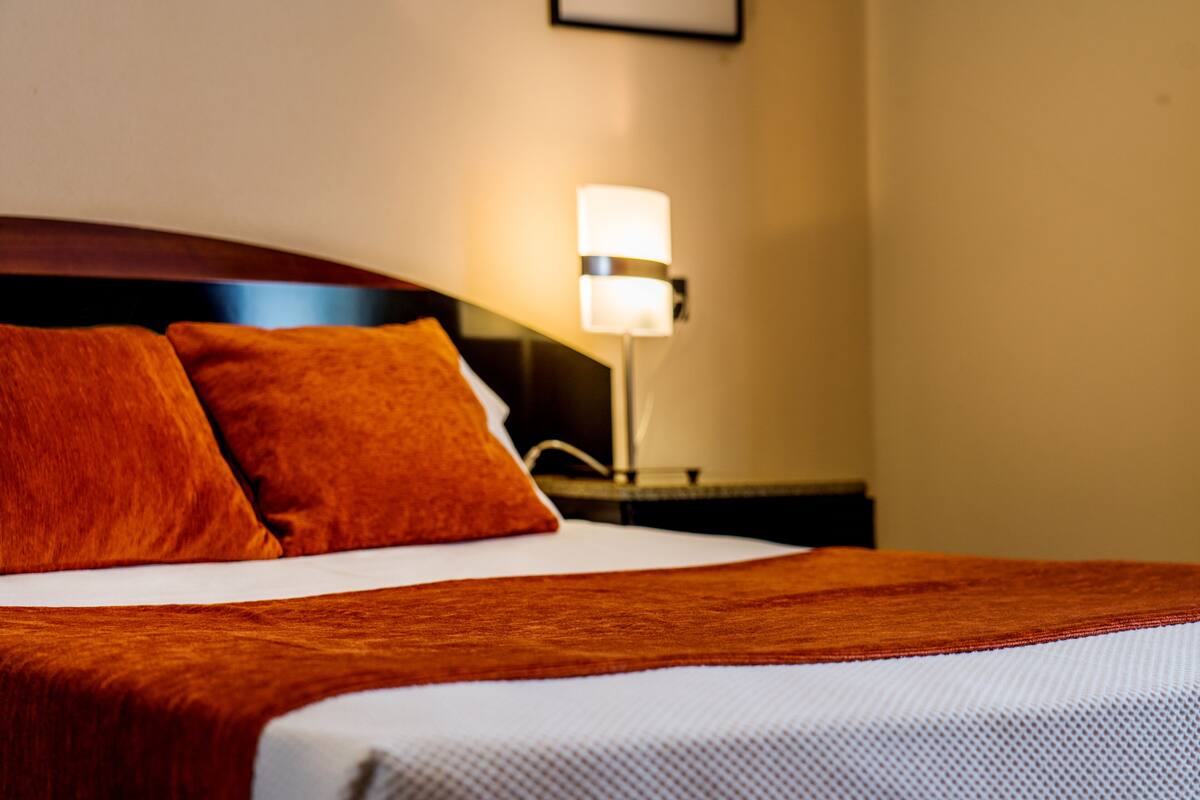 Cama bem arrumada com lençol branco e duas almofadas laranja, cabeceira de madeira e luminária de cabeceira com cúpula branca.