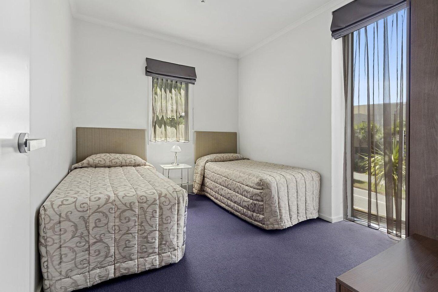 Quarto de hotel com duas camas de solteiro, uma mesinha, uma luminária e uma janela com cortinas.