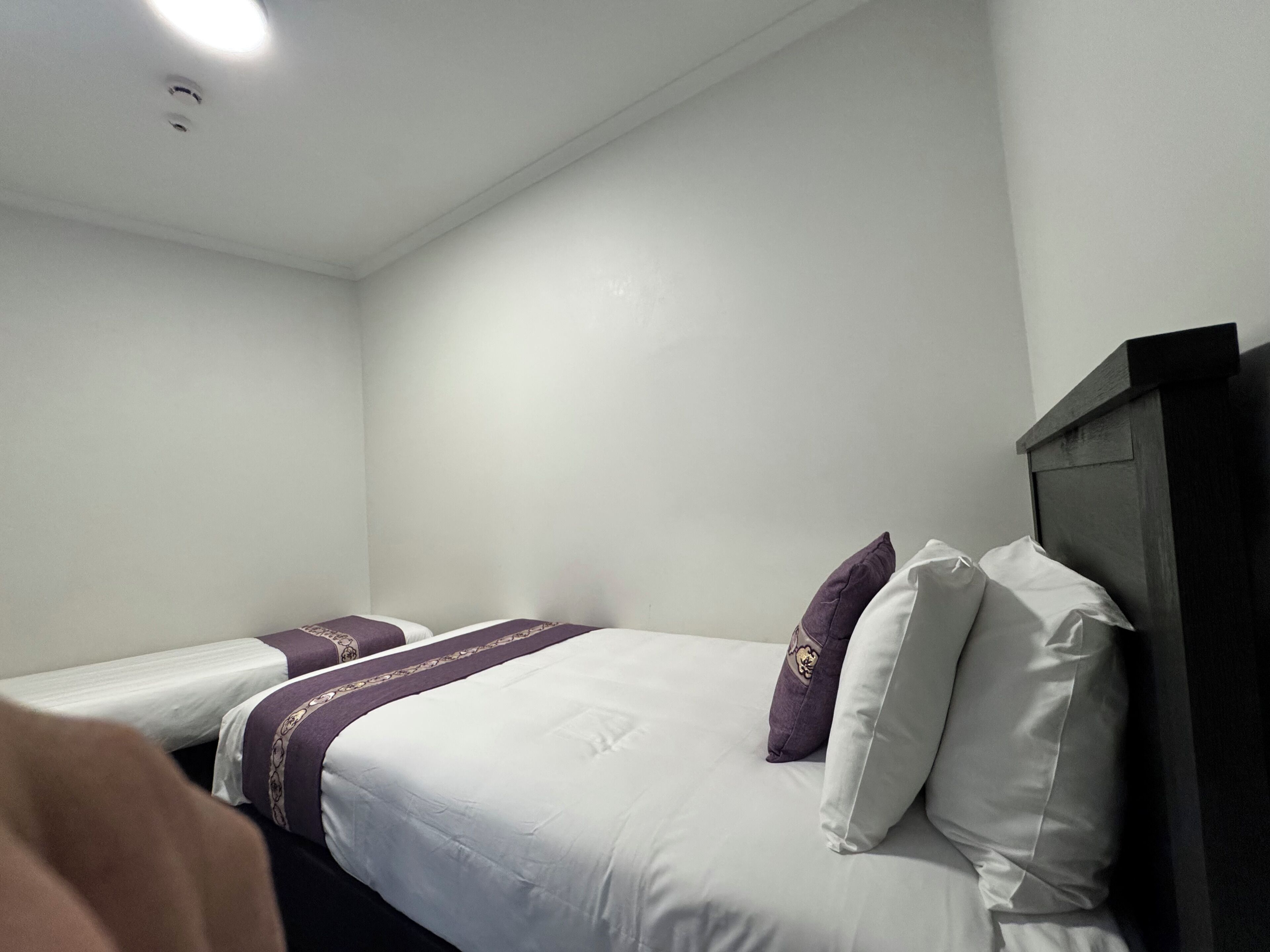Quarto pequeno com uma cama de solteiro, cabeceira de madeira, roupa de cama branca e um travesseiro roxo de destaque.