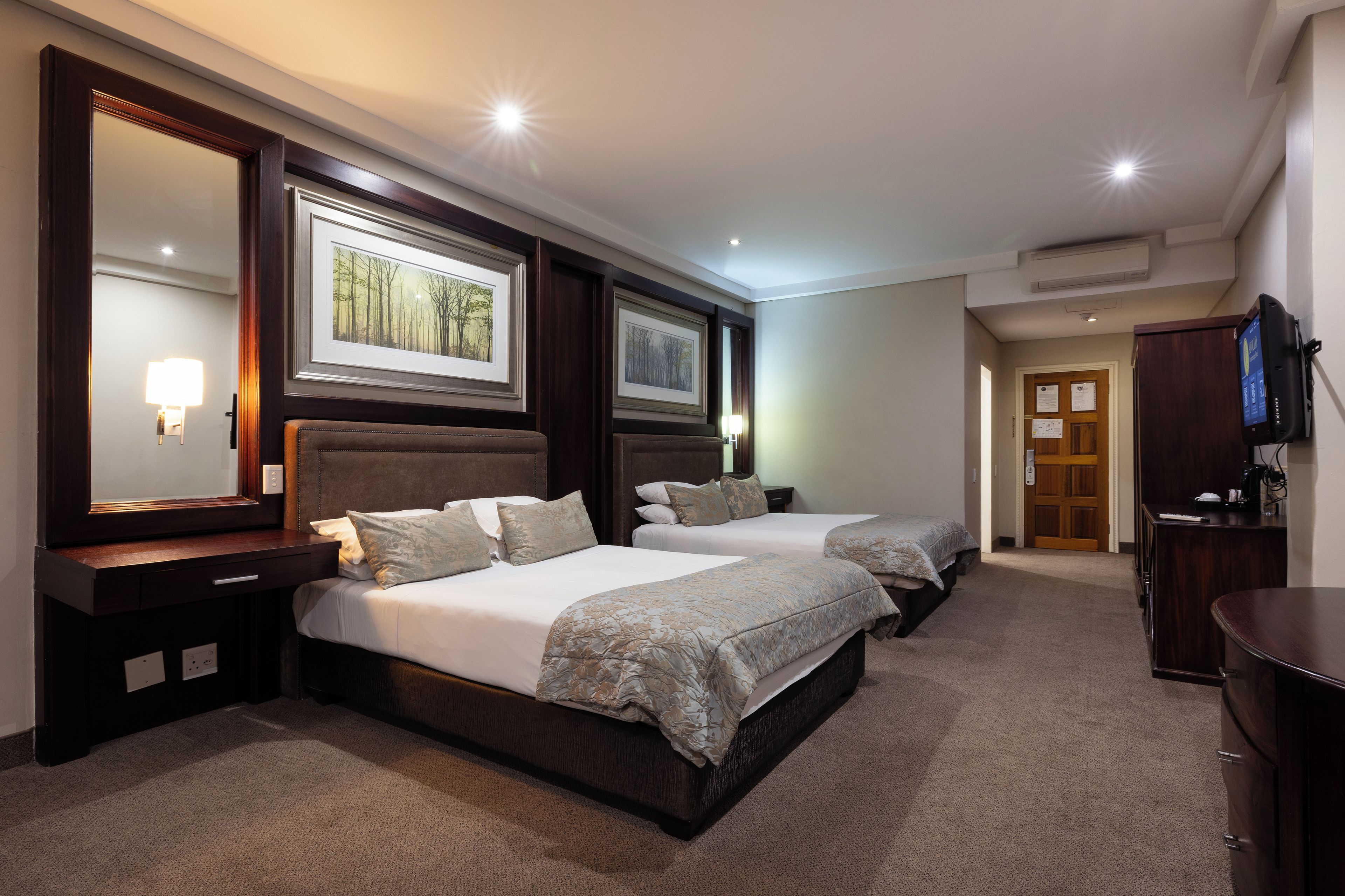 Quarto de hotel com duas camas, um espelho grande, quadros enquadrados, uma escrivaninha com televisão e uma porta.