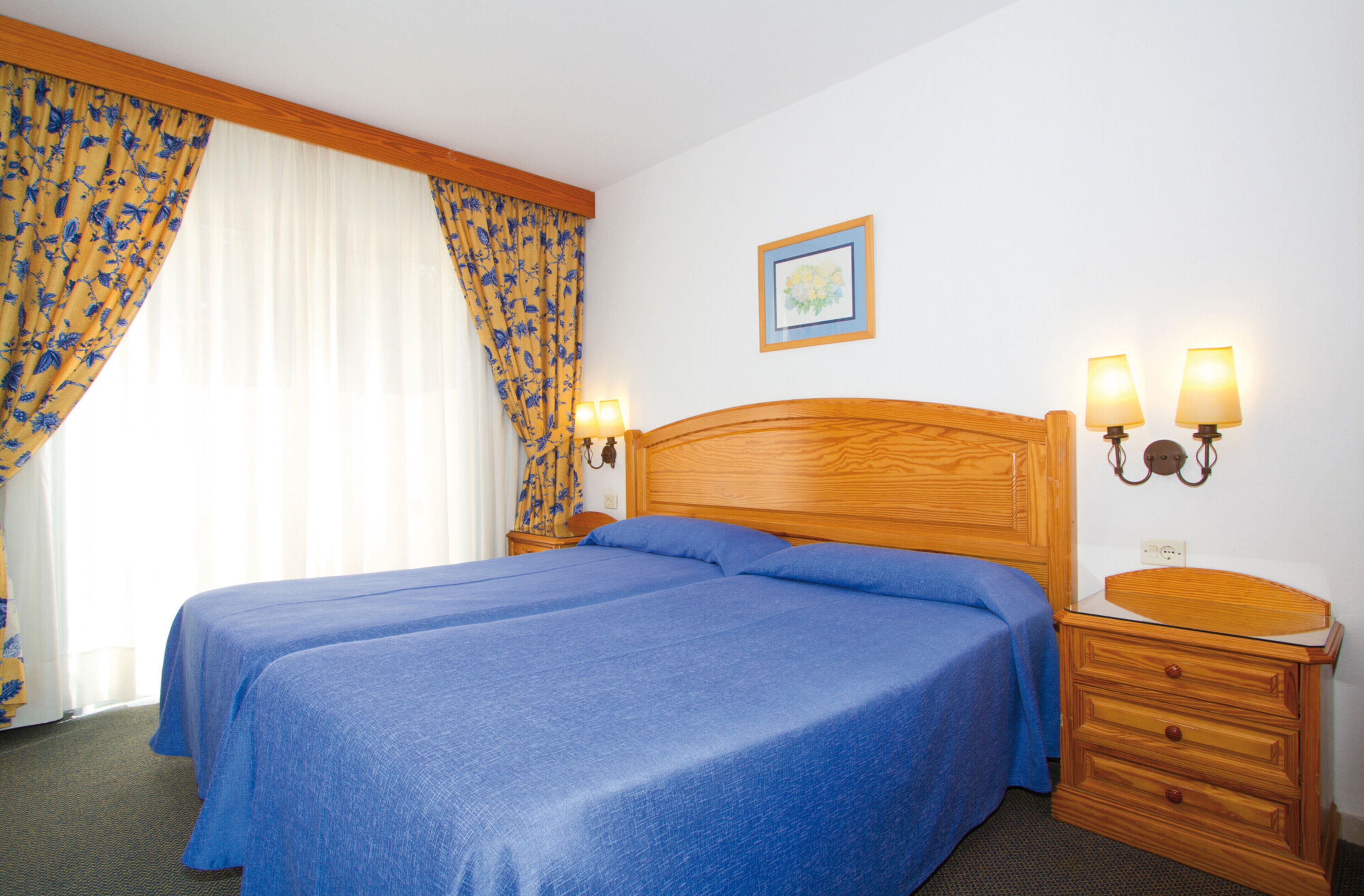 Uma cama de casal com cabeceira de madeira, duas luminárias de cabeceira e uma mesinha de noite com espelho.
