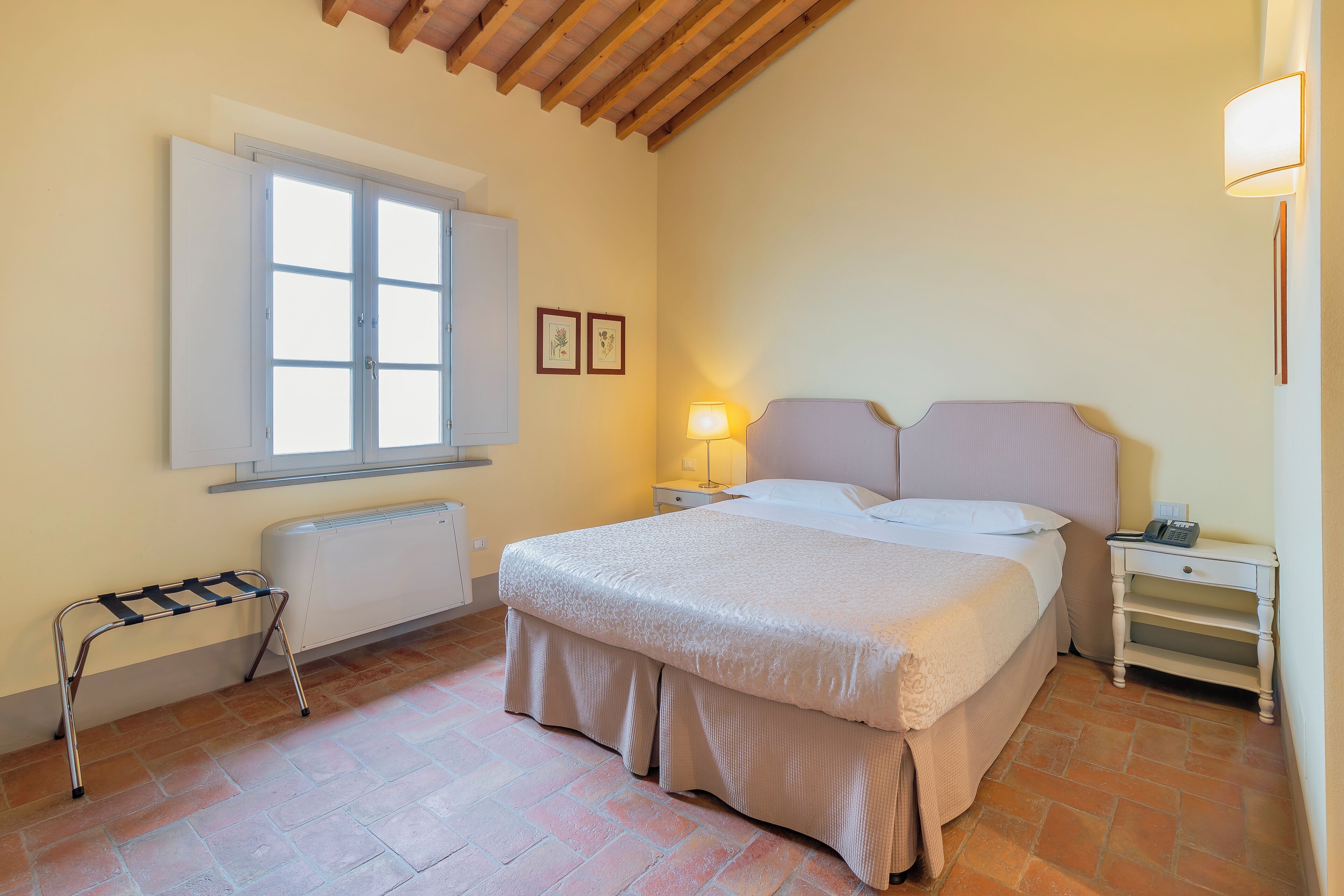 Quarto com cama, mesa de cabeceira, abajur, radiador e janela.